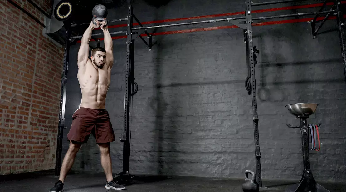 Mišičasti moški telovadi brez majice in izvaja vajo zamaha s kettlebell nad glavo