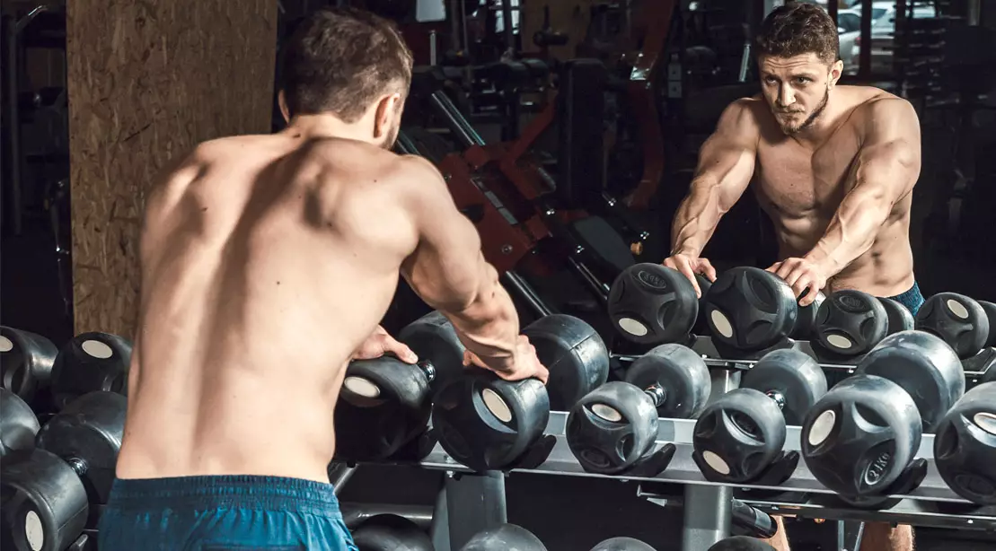 Muscular fitness model nga nagtan-aw sa salamin sunod sa dumbbell rack