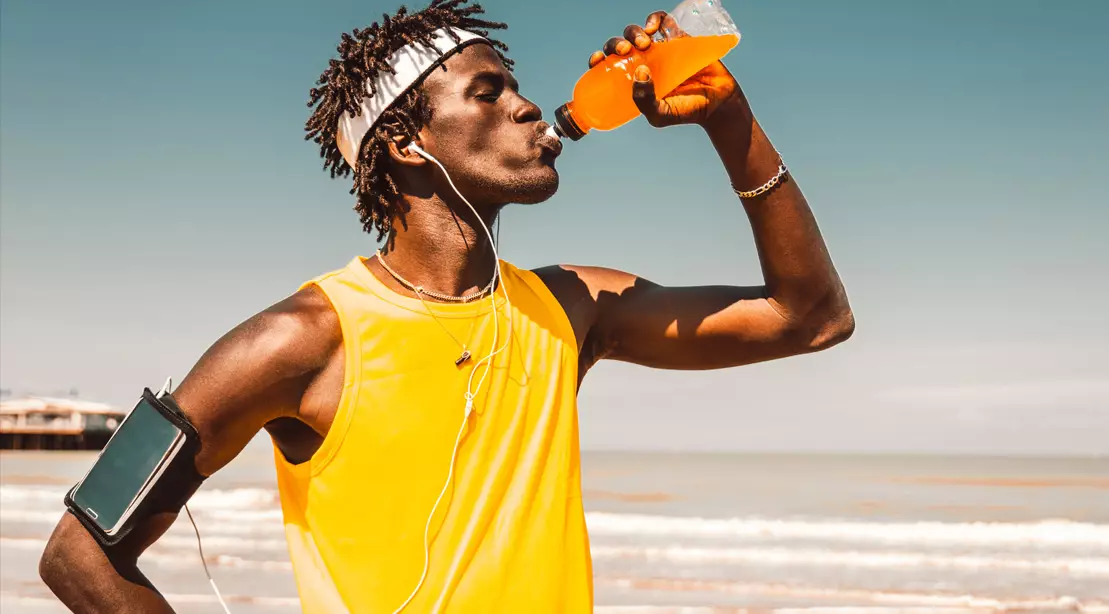 Fit runner na umiinom ng orange na sports drink at energy drink na may electrolytes habang tumatakbo sa beach para sa hydration