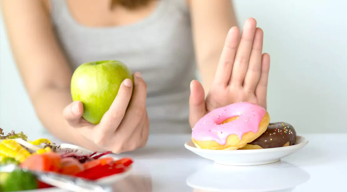 Vrouw eet een appel terwijl ze een bord met ongezond voedsel wegduwt.