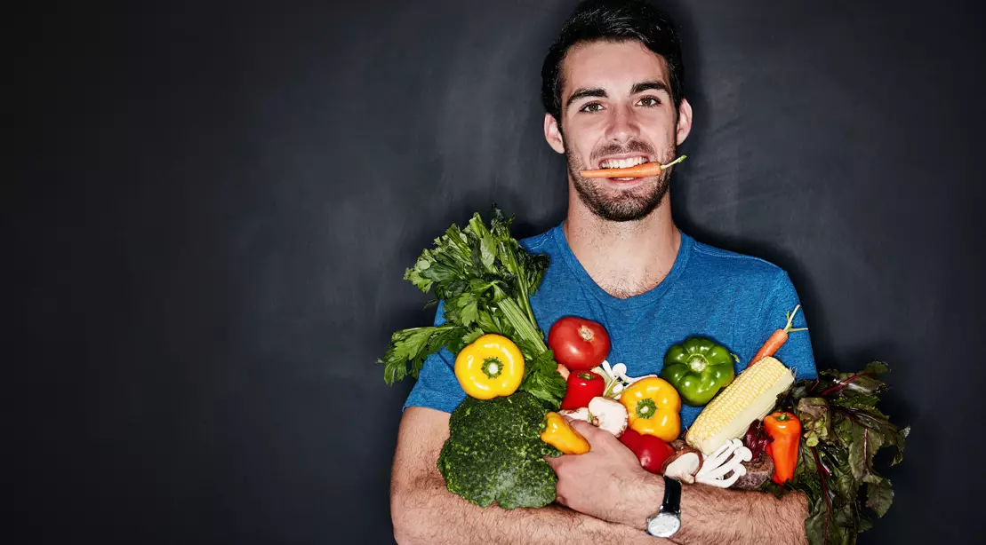Muž držící mrkev v ústech, zatímco drží pytel zeleniny a produktů pro svou volumetrickou dietu a nakupování na svém online obchodu s potravinami