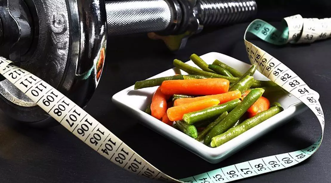 Bowl ng steamed vegetables sa tabi ng mga dumbbells at measuring tape