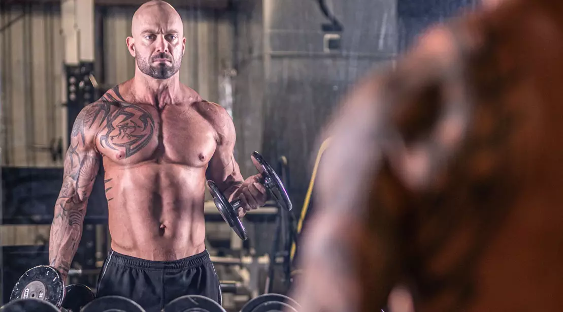 Justin Palionis realizando Dumbbell Curls na frente de um espelho no rack de halteres